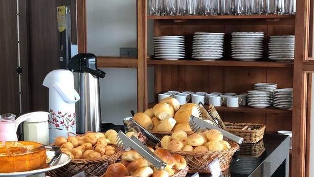 Buffet com diversos pães, doces e um bolo. Pratos e tigelas empilhados estão dispostos em prateleiras atrás do buffet.