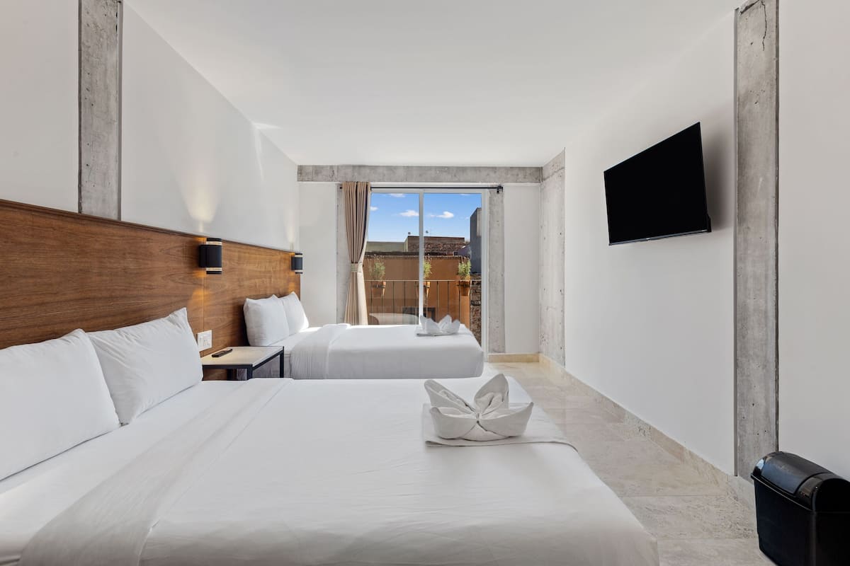 Quarto de hotel com duas camas, televisão de tela plana e uma janela grande com cortinas.
