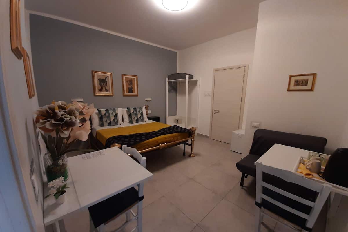 Quarto pequeno e bem organizado, com cama, mesa de cabeceira e uma cadeira. Há quadros nas paredes, uma mesinha com um vaso de flores e uma porta que dá para outro cômodo.