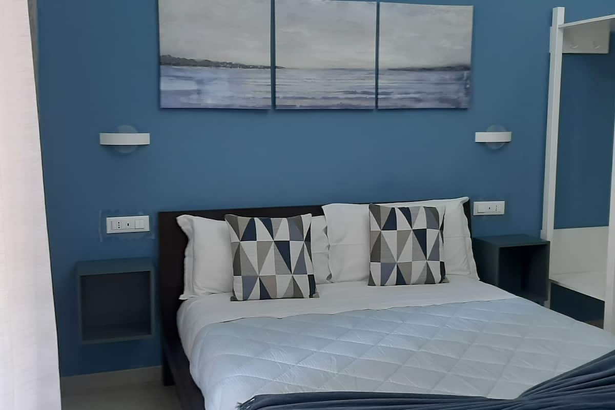 Quarto com cama, duas mesas de cabeceira, um criado-mudo, guarda-roupa e prateleira fixada na parede.