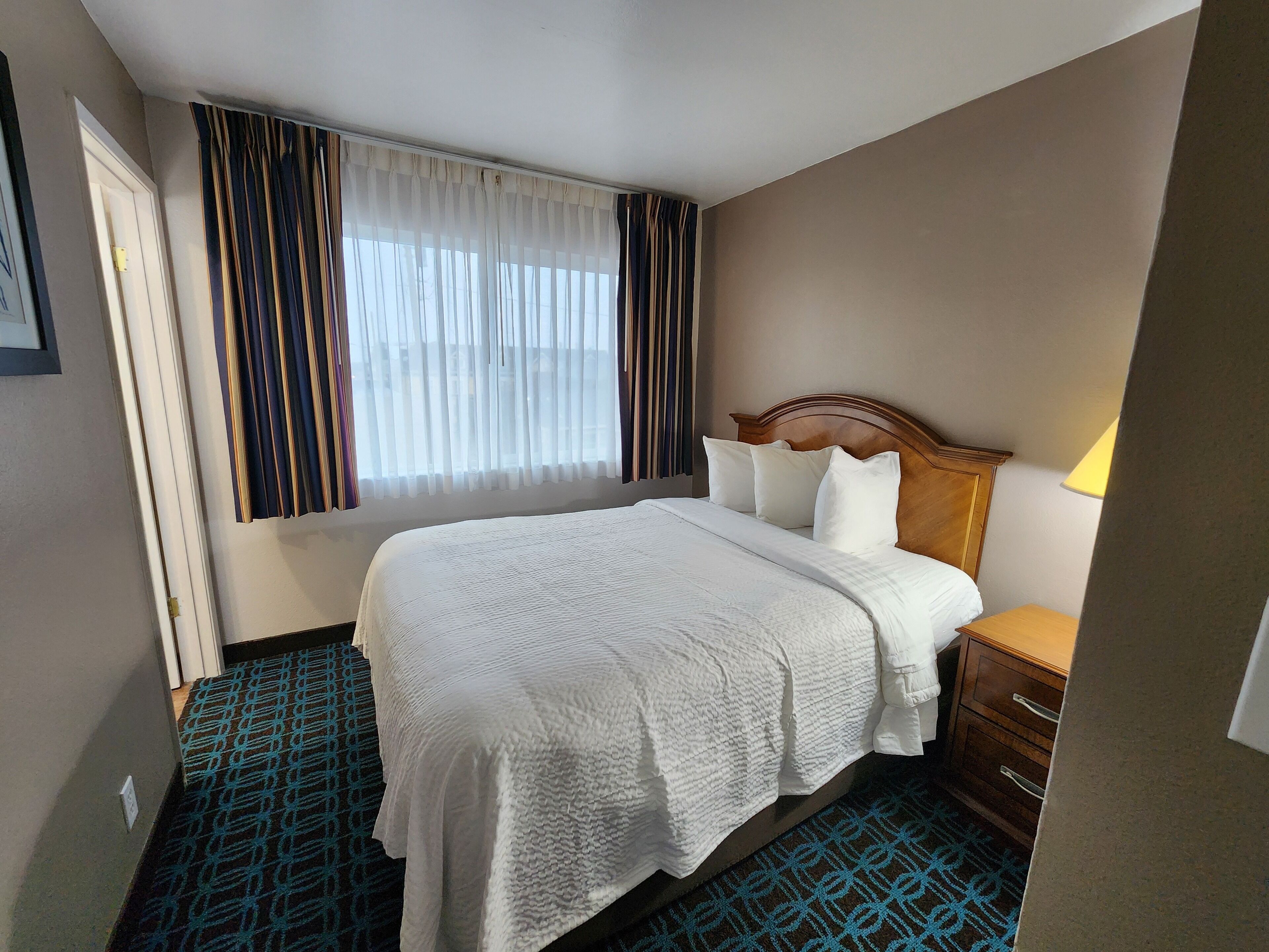 Quarto de hotel com uma cama grande, cabeceira de madeira, mesa de cabeceira com abajur e uma janela com cortinas.