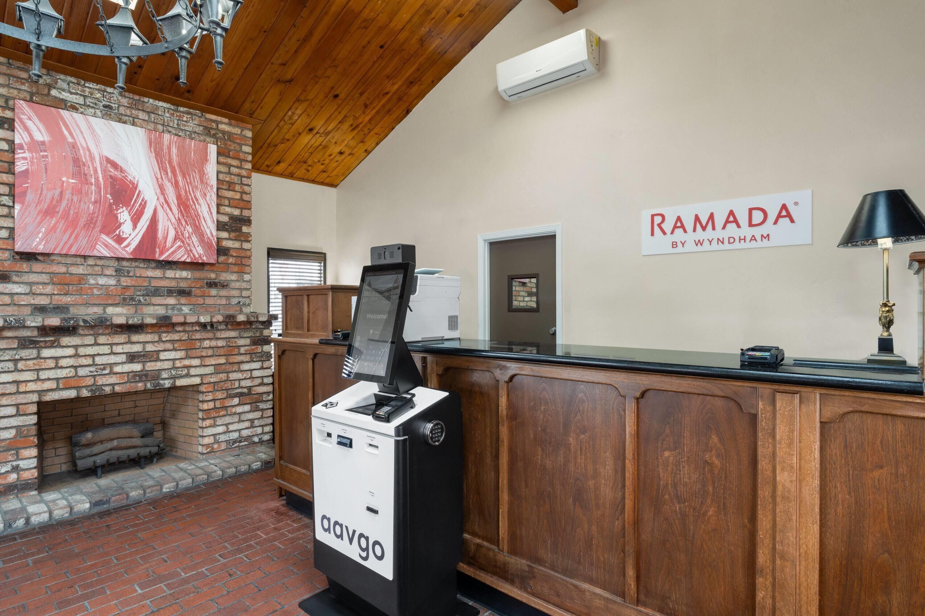 Uma área de recepção com lareira de tijolos, um quiosque digital, um balcão de madeira e uma placa com a inscrição 'RAMADA by Wyndham'.