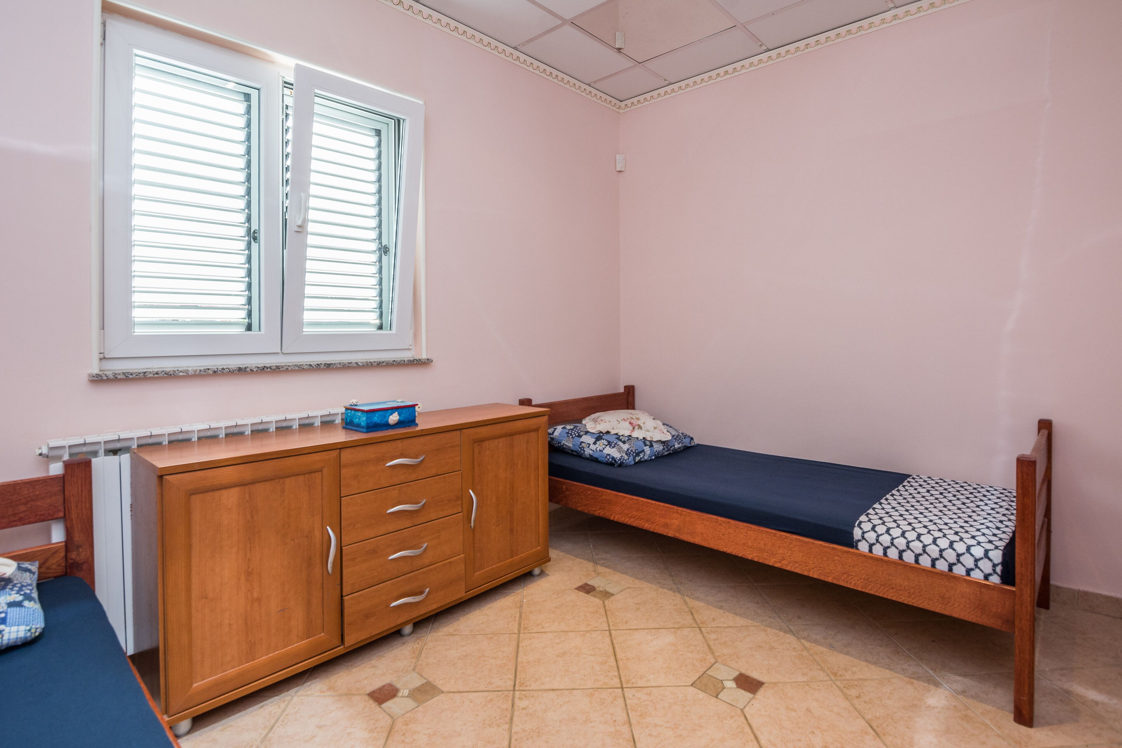 Quarto com uma cama de solteiro, guarda-roupa de madeira, radiador e janela com persianas.