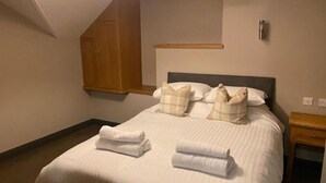 Cama bem arrumada com duas toalhas enroladas, um guarda-roupa de madeira e uma mesa de cabeceira com luminária.