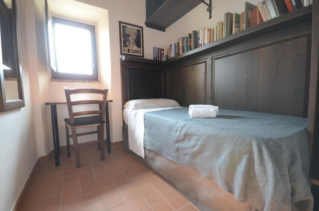 Um quarto pequeno e acolhedor com uma cama, uma cadeira, uma janela e uma estante.