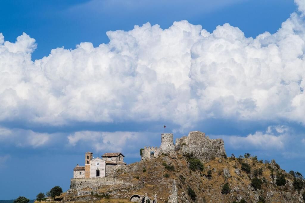 Um edifício histórico de pedra com uma torre de sino e uma estrutura fortificada numa colina, sob um céu parcialmente nublado.