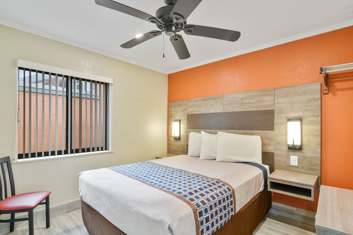 Quarto de hotel com cama, ventoinha no teto, janela com grades, cadeira e mesa de cabeceira.