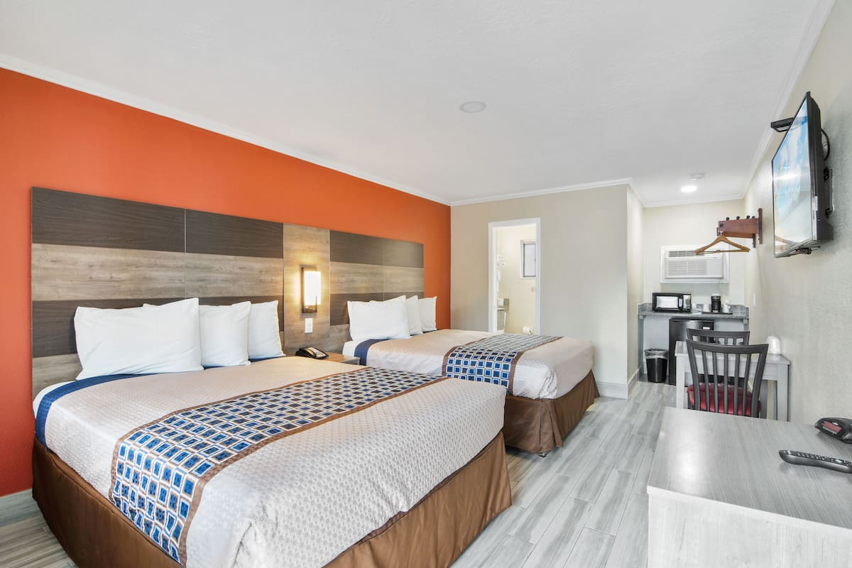 Quarto de hotel com duas camas, uma escrivaninha, uma cadeira, televisão e cozinha compacta.
