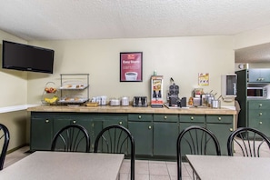 Uma cozinha compacta com máquina de café, torradeira e micro-ondas. Cadeiras e uma mesa estão em primeiro plano.
