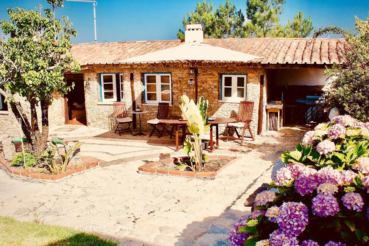 Uma casa de pedra com telhado em telhas, um pátio com mobiliário de exterior e um jardim com hortênsias.