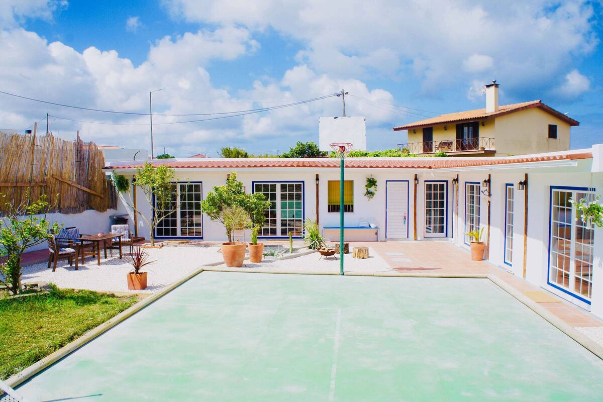 Um pátio com piscina, área de refeições com cadeiras e mesas, e um edifício com várias janelas e portas.