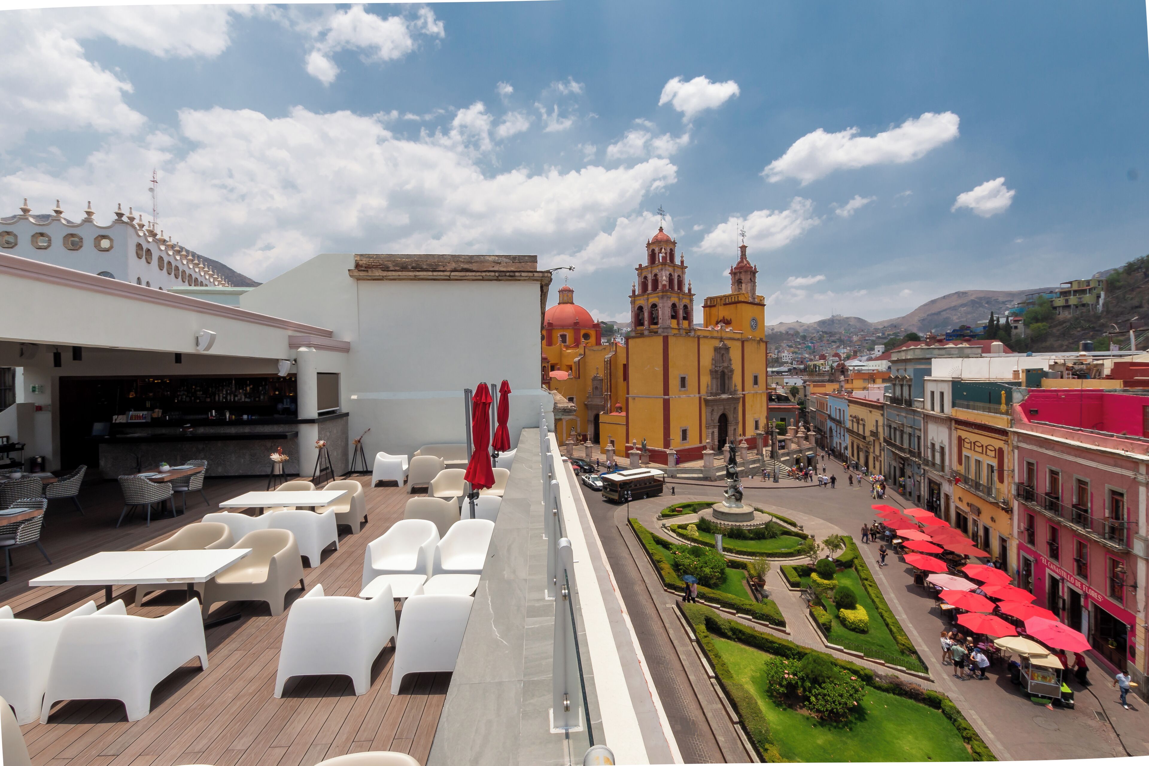 Um bar no terraço com cadeiras brancas e guarda-sóis vermelhos, uma área de bar e vista para uma igreja histórica com duas torres.