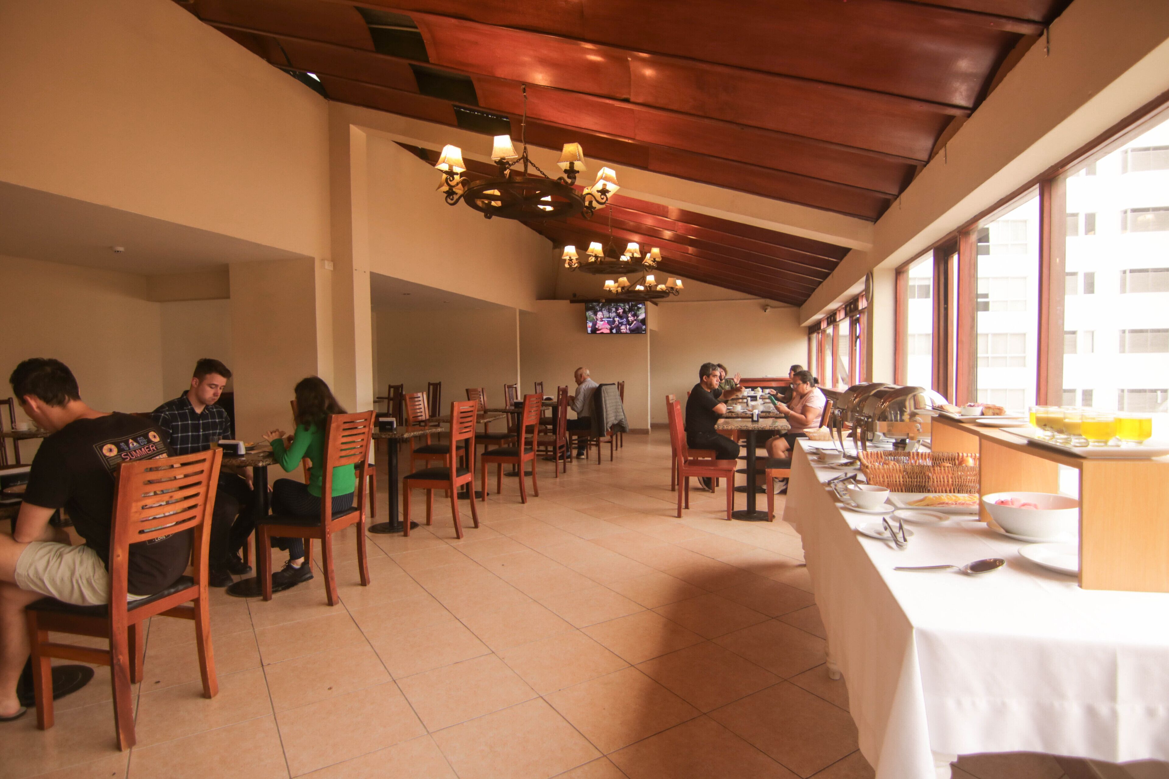 Restaurante com cadeiras e mesas de madeira, televisão na parede e grandes janelas que permitem entrada de luz natural.