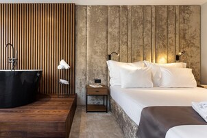 Quarto de hotel moderno com parede de ripas de madeira, banheira preta, mesa de cabeceira de madeira com abajur, cama com roupa de cama branca e um banco de couro marrom.