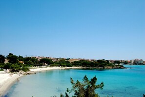 Uma praia costeira com águas turquesa cristalinas, areal arenoso e edifícios ao fundo.