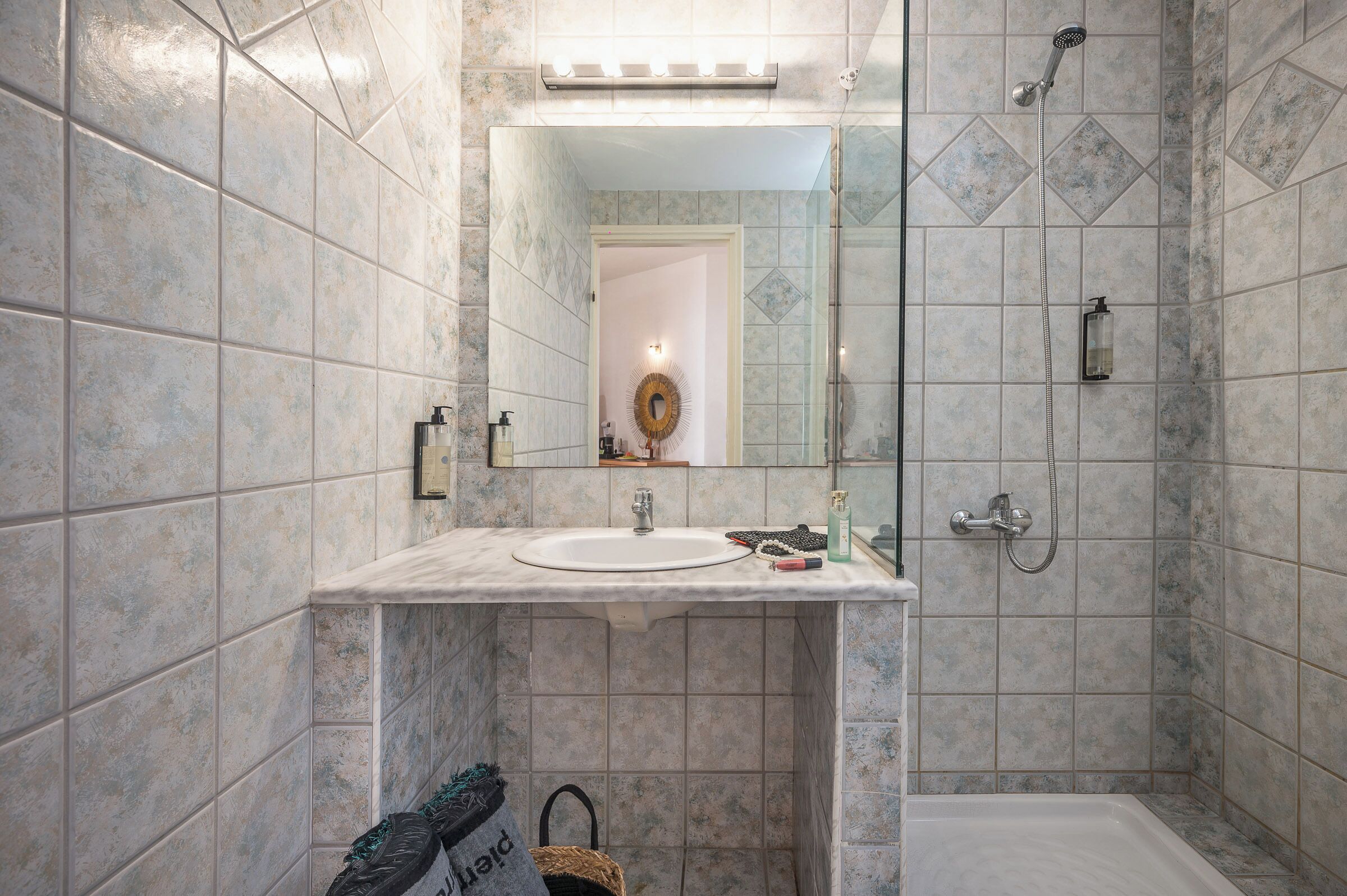 Casa de banho com lavatório, espelho, chuveiro e paredes em azulejo.