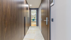 Um corredor com portas de madeira, piso ladrilhado e vista para um quarto com uma cadeira e uma mesa.