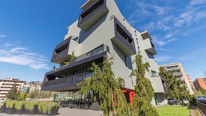 Um edifício moderno de vários andares, com varandas, terraço no telhado e vegetação ao redor.