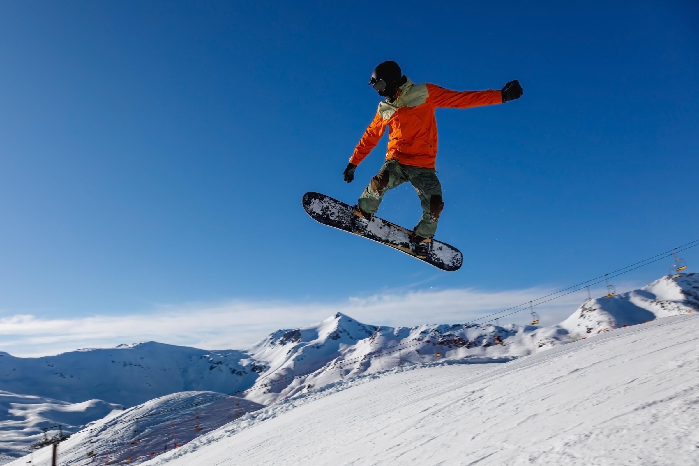 Um snowboarder no ar contra um céu azul límpido, com montanhas cobertas de neve ao fundo.
