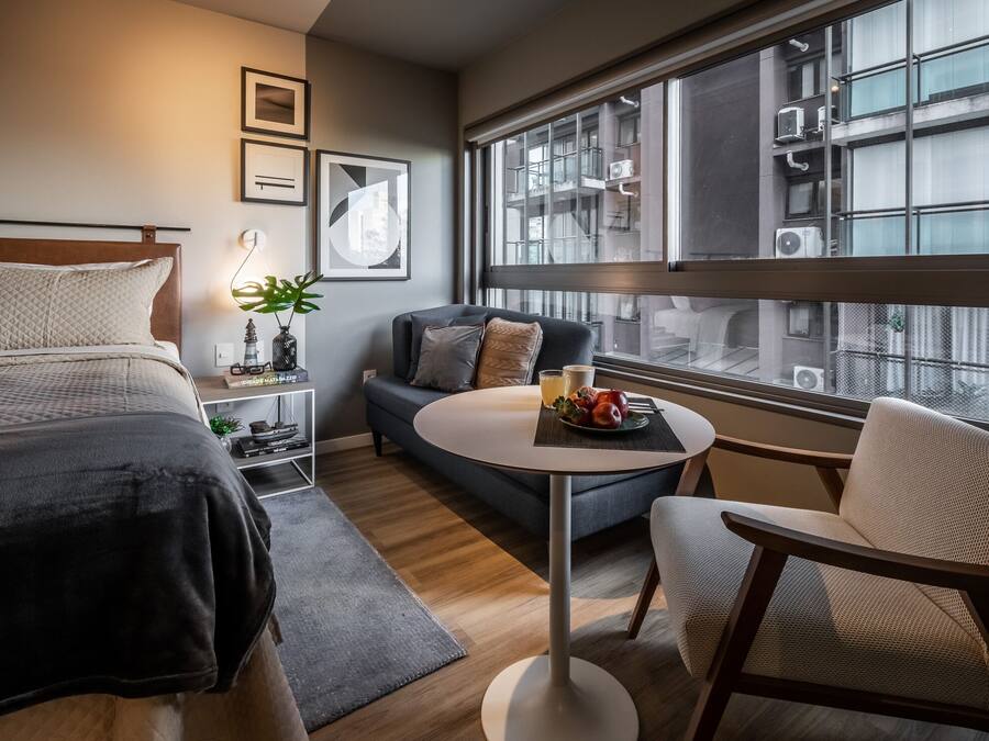Quarto de hotel moderno com cama, sofá, mesa redonda com frutas e uma janela grande com vista para a cidade.
