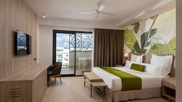 Quarto de hotel moderno com cama, escrivaninha, cadeira, banqueta, televisão, ventilador de teto e varanda com vista.