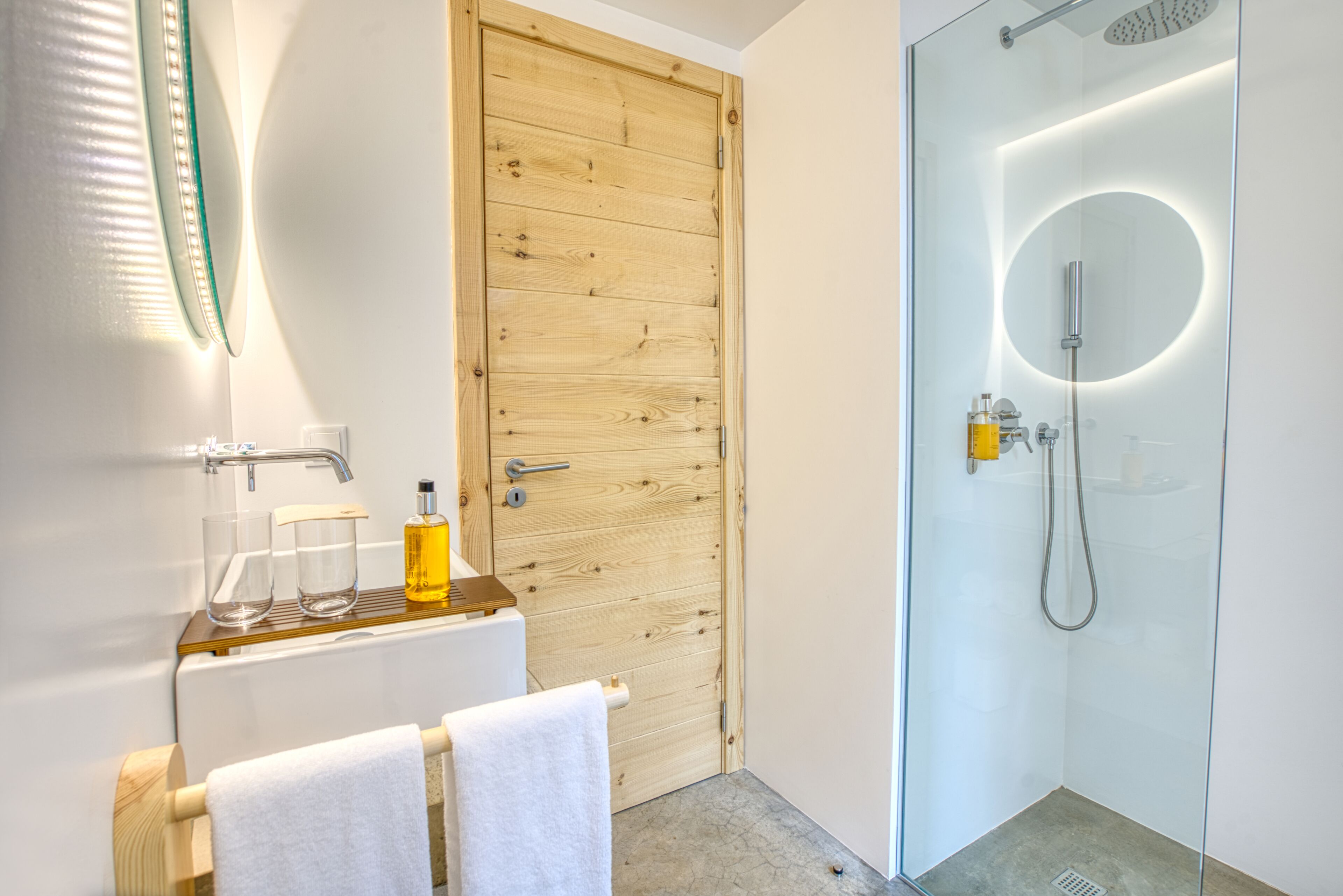 Uma casa de banho moderna com porta de madeira, cabine de chuveiro em vidro e lavatório branco com dois copos e uma garrafa.