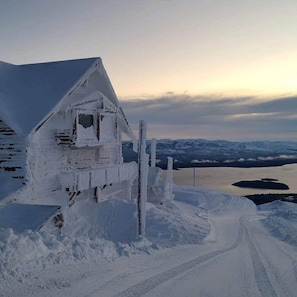 Um edifício coberto de neve com icicles, céu claro e vista distante de montanhas e um lago.