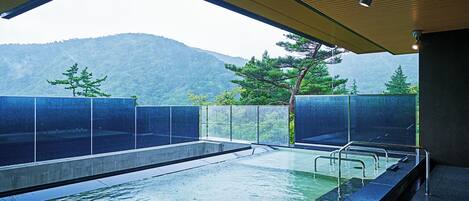 Piscina interior moderna com paredes de vidro transparente, vista para uma montanha arborizada e teto de madeira.