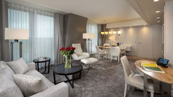 Quarto de hotel moderno com sofá, mesa de jantar e mesa de centro. Há duas luminárias e um vaso com flores sobre a mesa de centro.