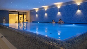 Uma piscina interna com azulejos de mosaico azul, uma parede de vidro e luminárias fixadas na parede.
