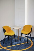 Duas cadeiras amarelas com estrutura preta e uma pequena mesa branca com base dourada, dispostas sobre um tapete azul com linhas amarelas.