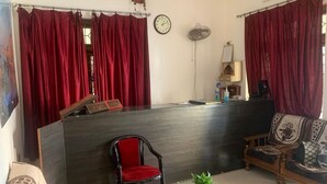 Uma área de recepção com um balcão escuro, cortinas vermelhas, um relógio, um ventilador e uma pequena planta.