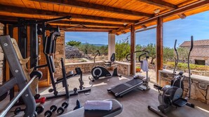 Ginásio ao ar livre coberto, com equipamentos de exercício, parede de pedra e vista para o campo.