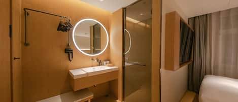 Um banheiro de hotel moderno com um espelho redondo iluminado, chuveiro e uma cama.