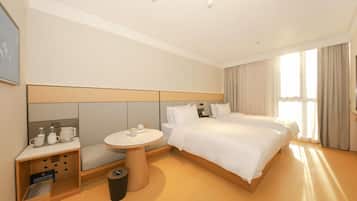 Quarto de hotel com uma cama grande, uma mesa redonda com uma cadeira, um pequeno espaço de estar com um banco e uma lixeira.