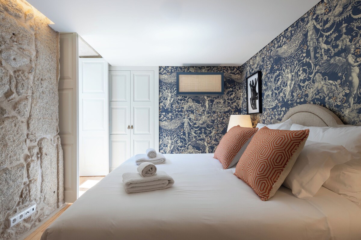 Quarto com parede de pedra, cama com roupa de cama branca, almofadas estampadas e uma obra de arte pendurada na parede.