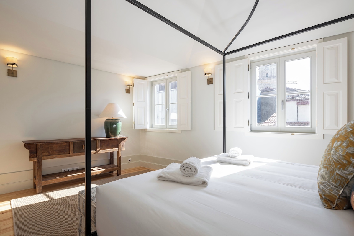 Quarto com cama grande, um banquinho de madeira, uma luminária e uma janela com persianas.