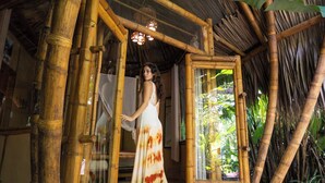 Estrutura tradicional de bambu com telhado de palha, um banco de madeira e porta de vidro que dá para o interior.