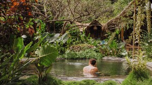 Um homem em uma piscina natural rodeado por vegetação exuberante e uma cabana rústica.