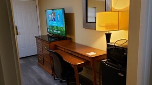 Quarto de hotel com escrivaninha de madeira, televisão, luminária e lixeira.