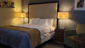 Cama bem arrumada com lençóis brancos e manta estampada, acompanhada por duas mesinhas de cabeceira com abajures, uma poltrona estofada verde e um quadro na parede.