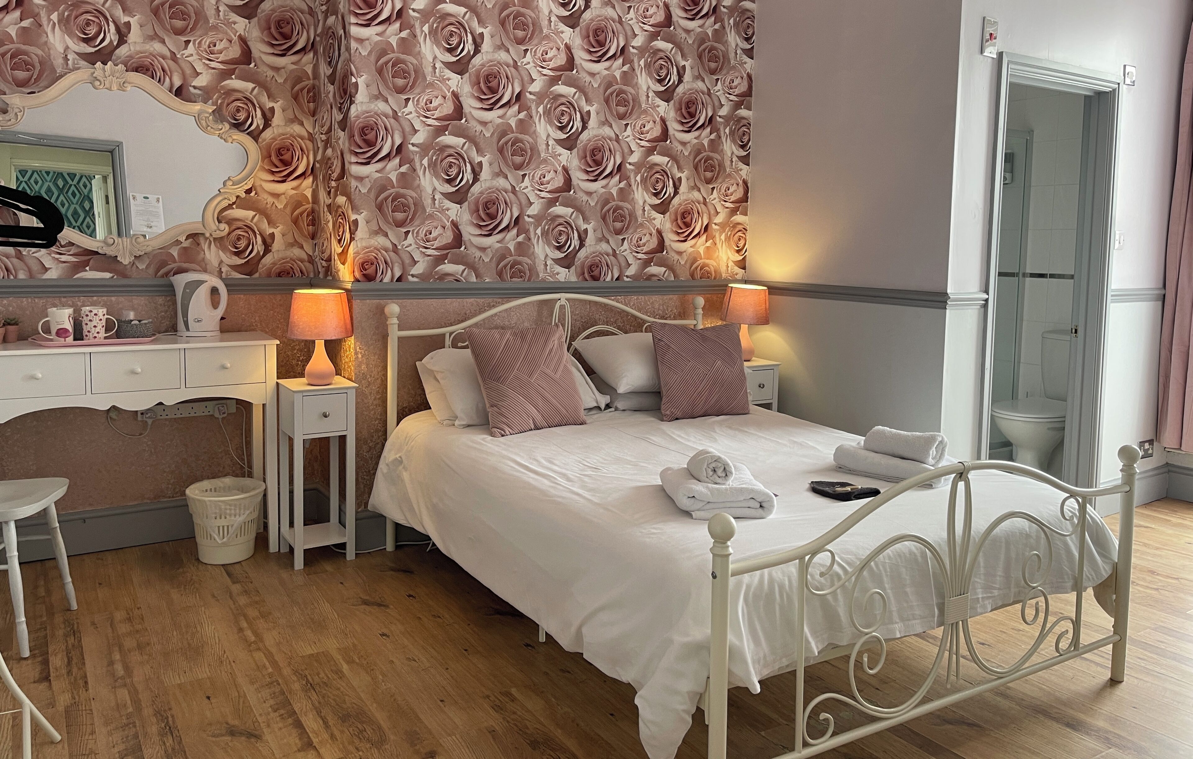 Quarto com cama branca, papel de parede estampado em rosa, espelho, cômoda e mesa de cabeceira com abajur.