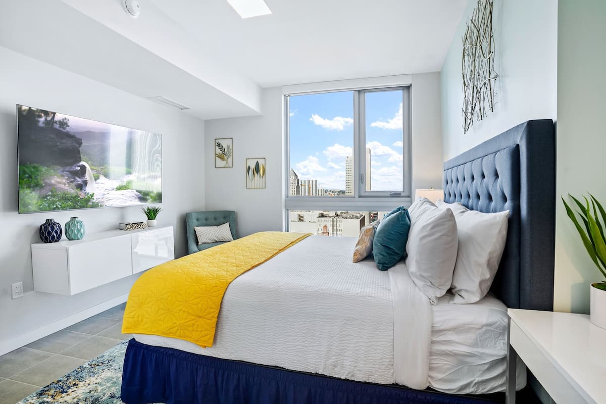 Quarto moderno com cama grande, vista para a cidade, televisão montada e um console branco.