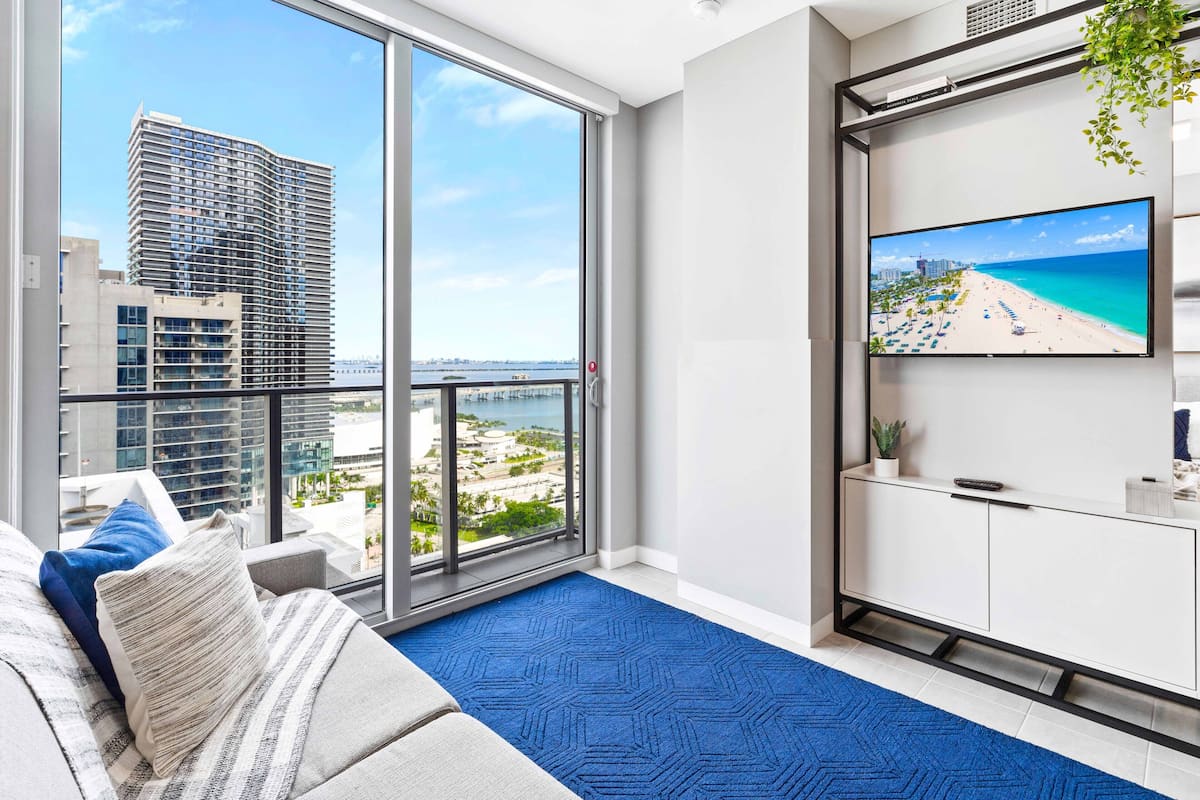 Quarto de hotel moderno com uma grande janela que oferece vista da cidade, um tapete azul estampado, um sofá com almofadas, uma TV na parede e uma pequena planta no aparador.