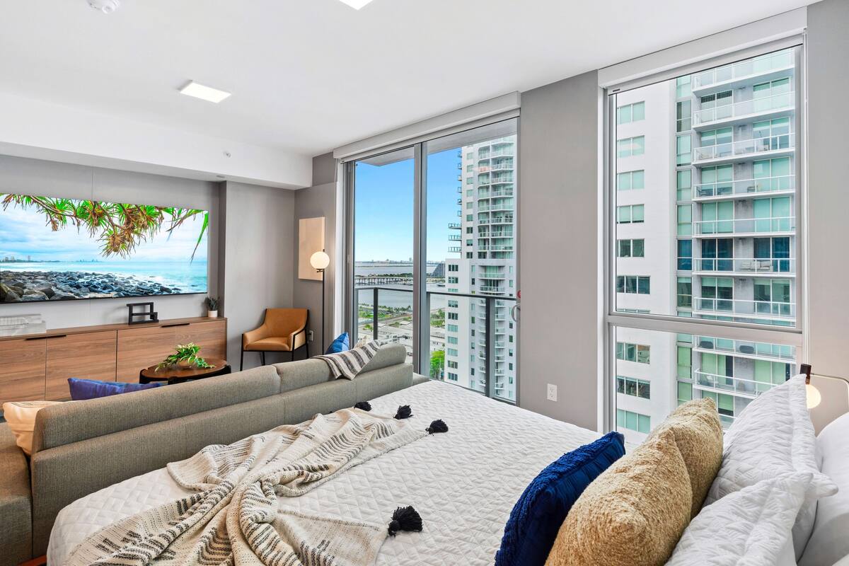 Quarto de hotel moderno com uma cama grande, um sofá, uma mesa de trabalho e vista da cidade por grandes janelas.
