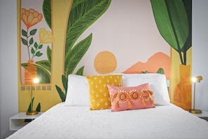 Uma cama com roupa de cama branca, duas almofadas estampadas, uma mesa de cabeceira com abajur e um mural na parede com motivos de plantas e sol.
