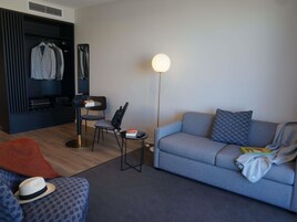 Uma sala de estar moderna com um sofá cinzento, um abajur redondo, uma pequena mesa com uma cadeira, um guarda-roupa com roupas penduradas e um banquinho estampado.