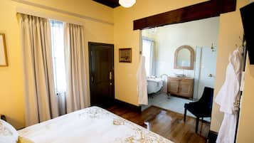 Quarto com cama grande, piso de madeira, porta e vista para um banheiro com banheira e espelho.