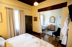 Quarto com cama grande, piso de madeira, porta e vista para um banheiro com banheira e espelho.