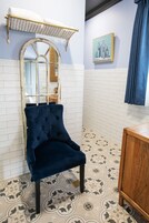 Um banheiro com uma cadeira de veludo azul, um armário de madeira, um espelho com moldura decorativa e um piso de azulejos com padrão floral.